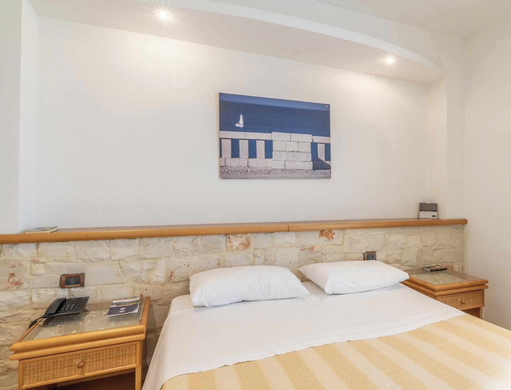 Quarto de hotel com cama, duas mesas de cabeceira, criado-mudo, telefone fixado na parede e uma moldura com imagem.