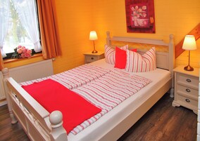 Cama bem arrumada com manta vermelha e roupa de cama listrada, duas luminárias de cabeceira, mesa de cabeceira e janela com cortinas.