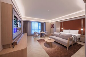 Quarto de hotel moderno com uma televisão de tela grande, um sofá, uma cama, uma mesa de centro e uma pequena mesa redonda com cadeiras.