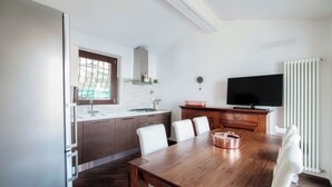 Cozinha moderna com área de refeições, destacando uma mesa de madeira, cadeiras brancas, uma televisão e uma janela ampla.