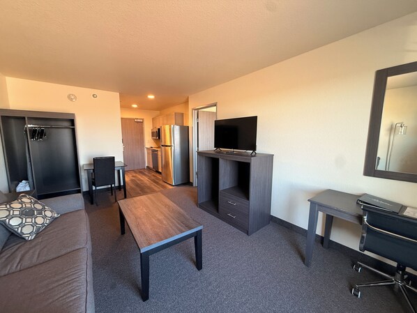 Quarto de hotel compacto com sofá, mesa pequena, televisão em móvel, escrivaninha e guarda-roupa.