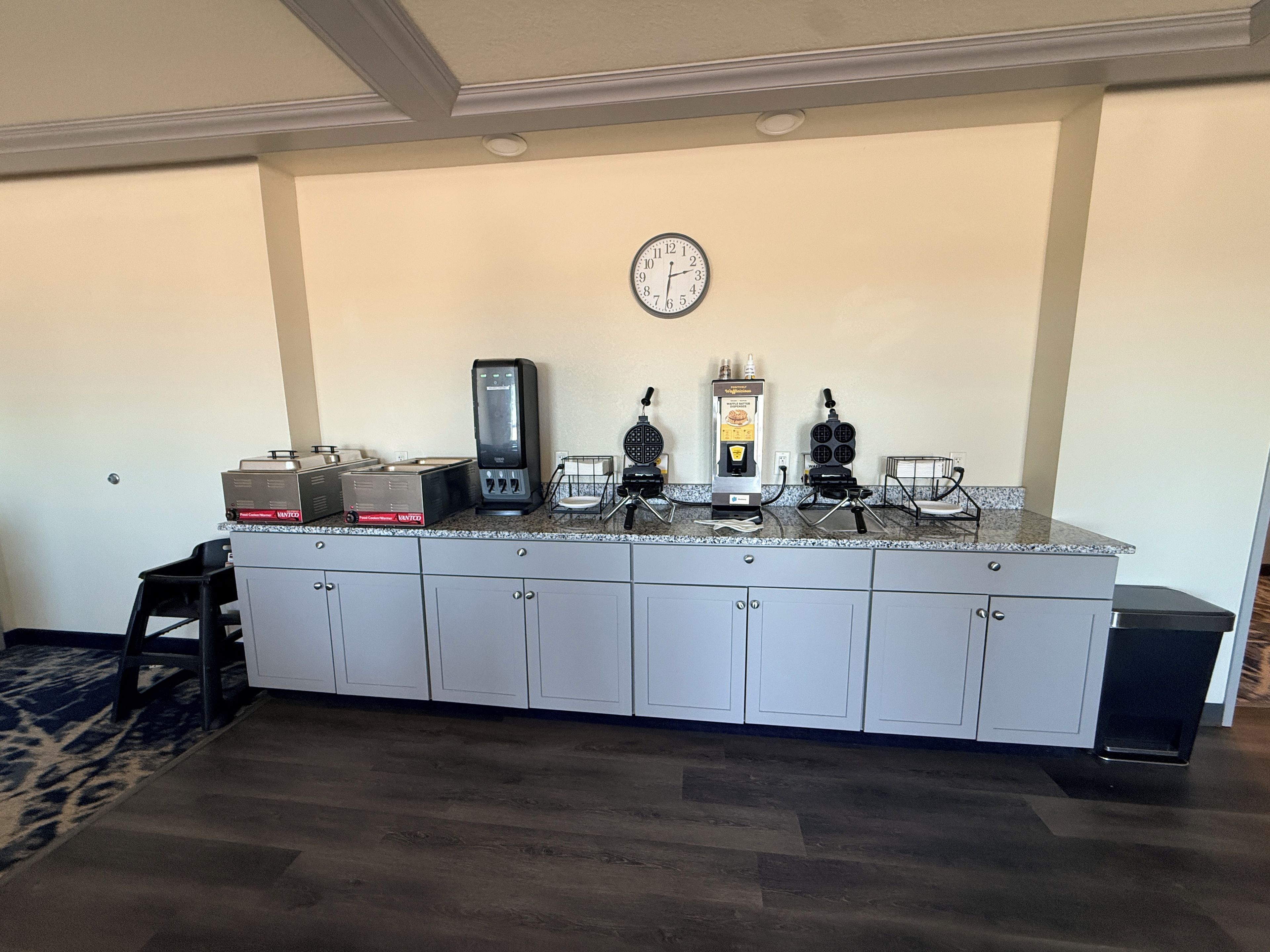 Quarto de hotel com cozinha compacta, incluindo máquina de café, aparelho para waffles e um relógio na parede.