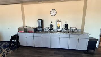 Quarto de hotel com cozinha compacta, incluindo máquina de café, aparelho para waffles e um relógio na parede.