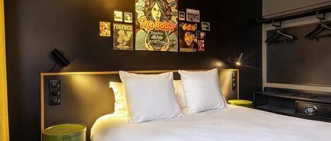 Quarto de hotel com uma cama grande, cabeceira, TV fixada na parede e um cesto de lixo verde.