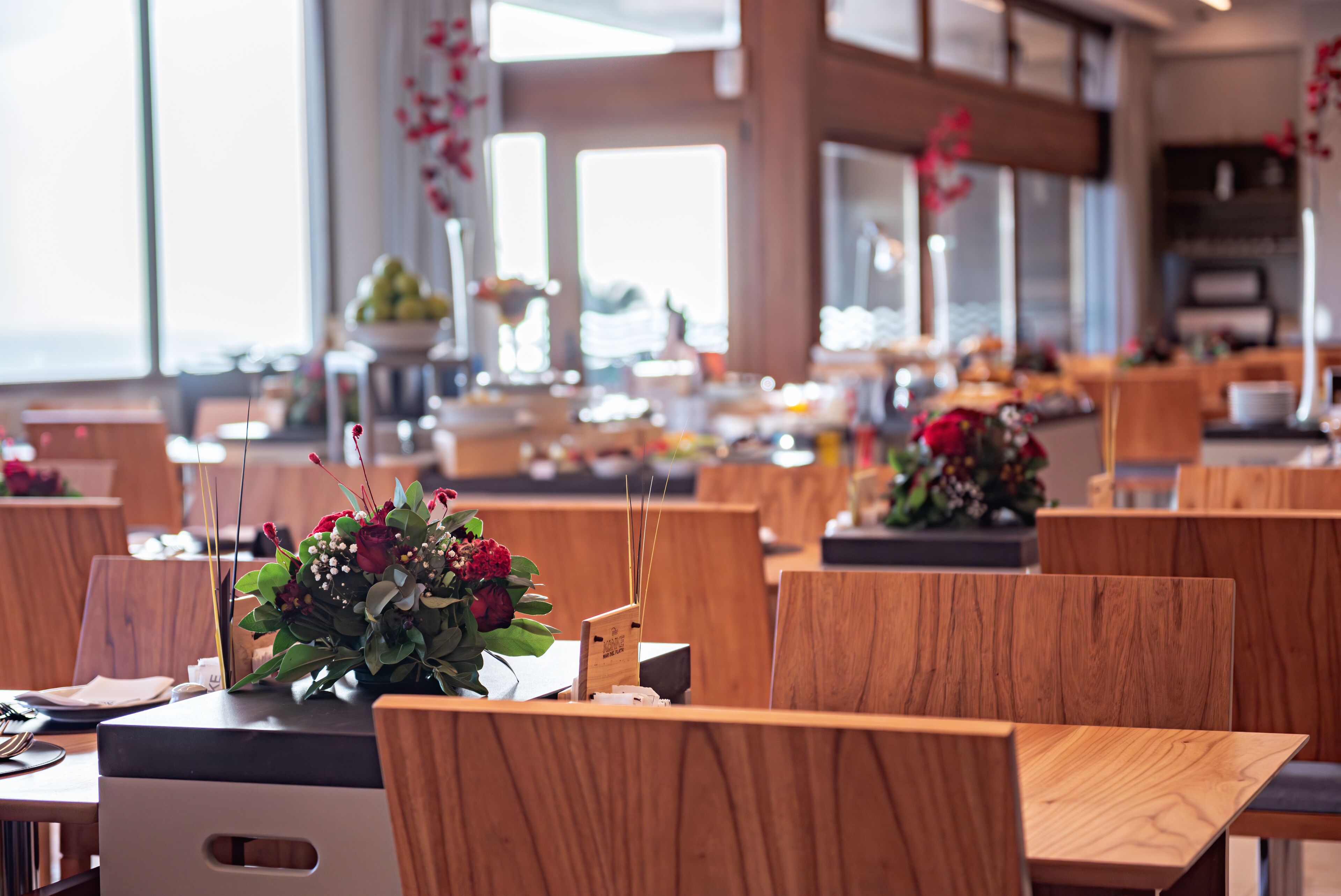 Um restaurante com mesas e cadeiras de madeira, centros de mesa florais e grandes janelas que permitem a entrada de luz natural.