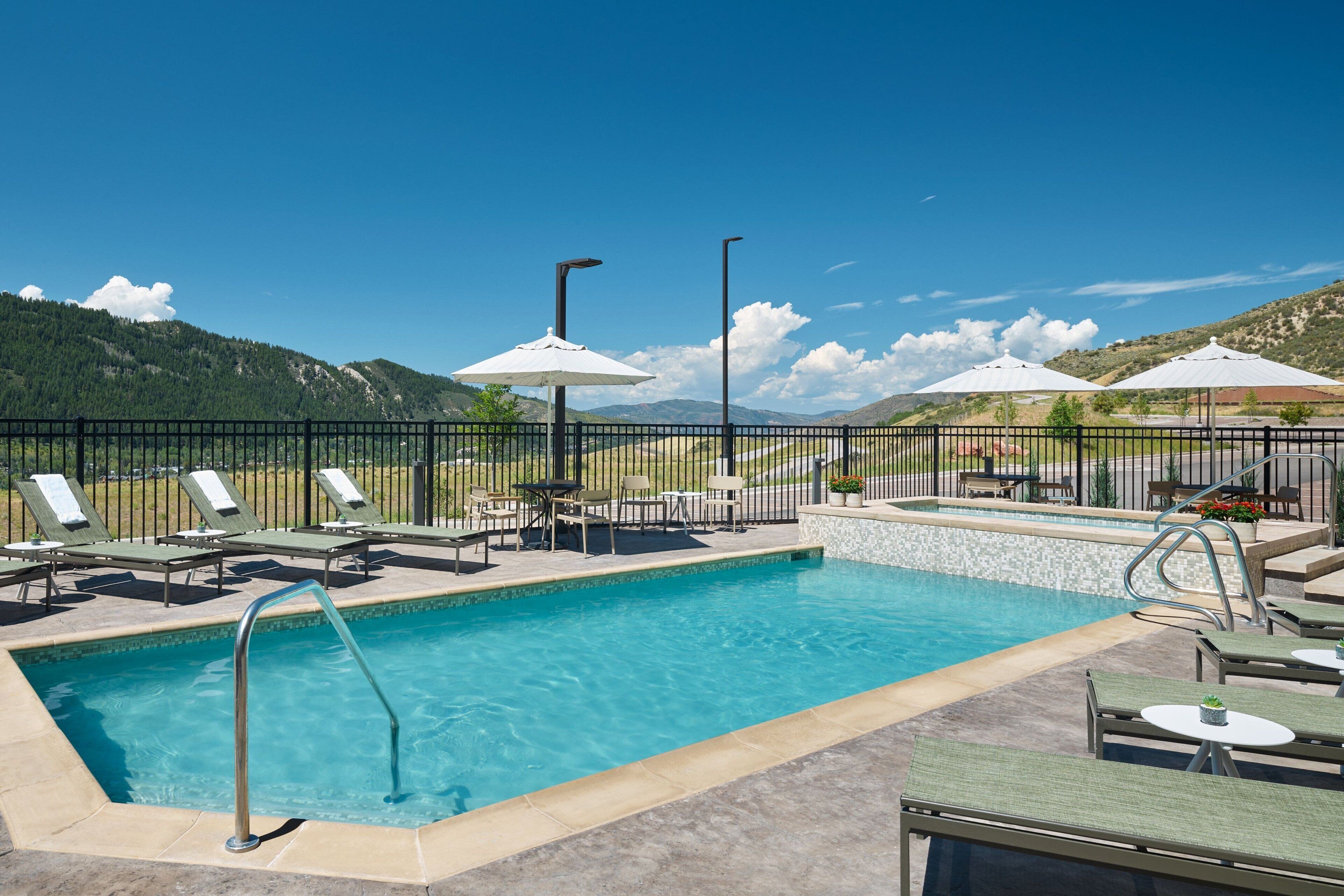 Piscina com espreguiçadeiras e guarda-sóis, cercada por uma vedação e montanhas ao fundo.