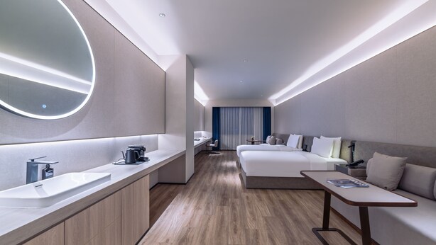 Quarto de hotel moderno com uma cama grande, mesa de cabeceira, escrivaninha, espelho redondo e banheiro com pia e vaso sanitário.