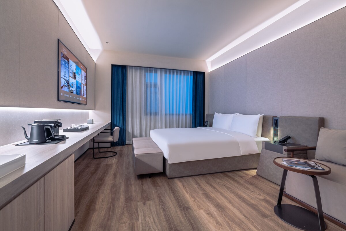 Quarto de hotel moderno com cama, mesa de cabeceira, pequena escrivaninha com cafeteira, cadeira e vista da cidade através de grandes janelas.