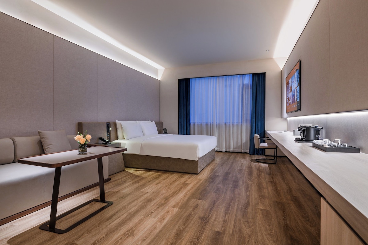Quarto de hotel moderno com cama, sofá, mesa pequena, cozinha compacta e uma janela grande com cortinas.