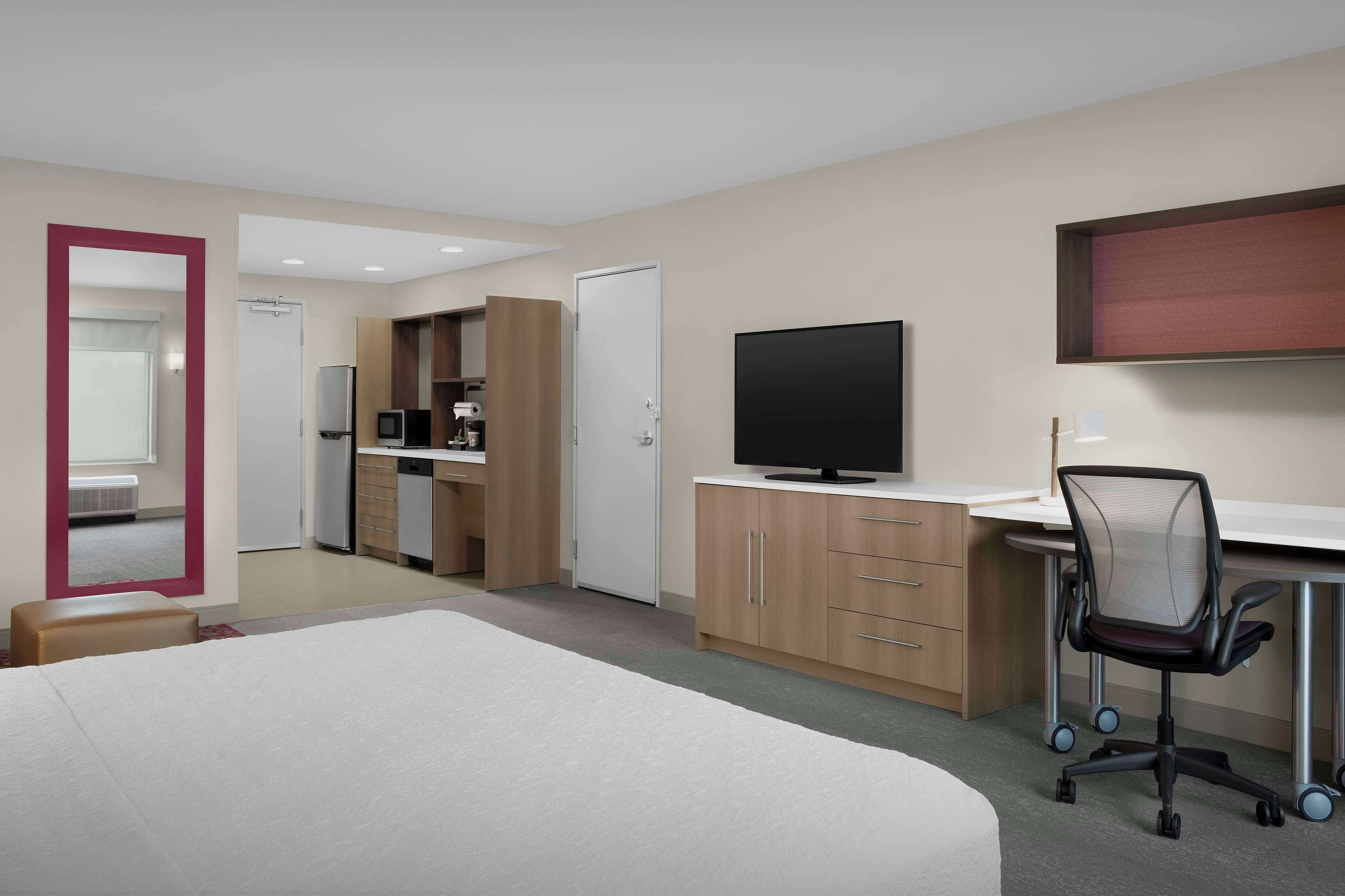 Quarto de hotel com cama, escrivaninha, cadeira, televisão e cozinha compacta.