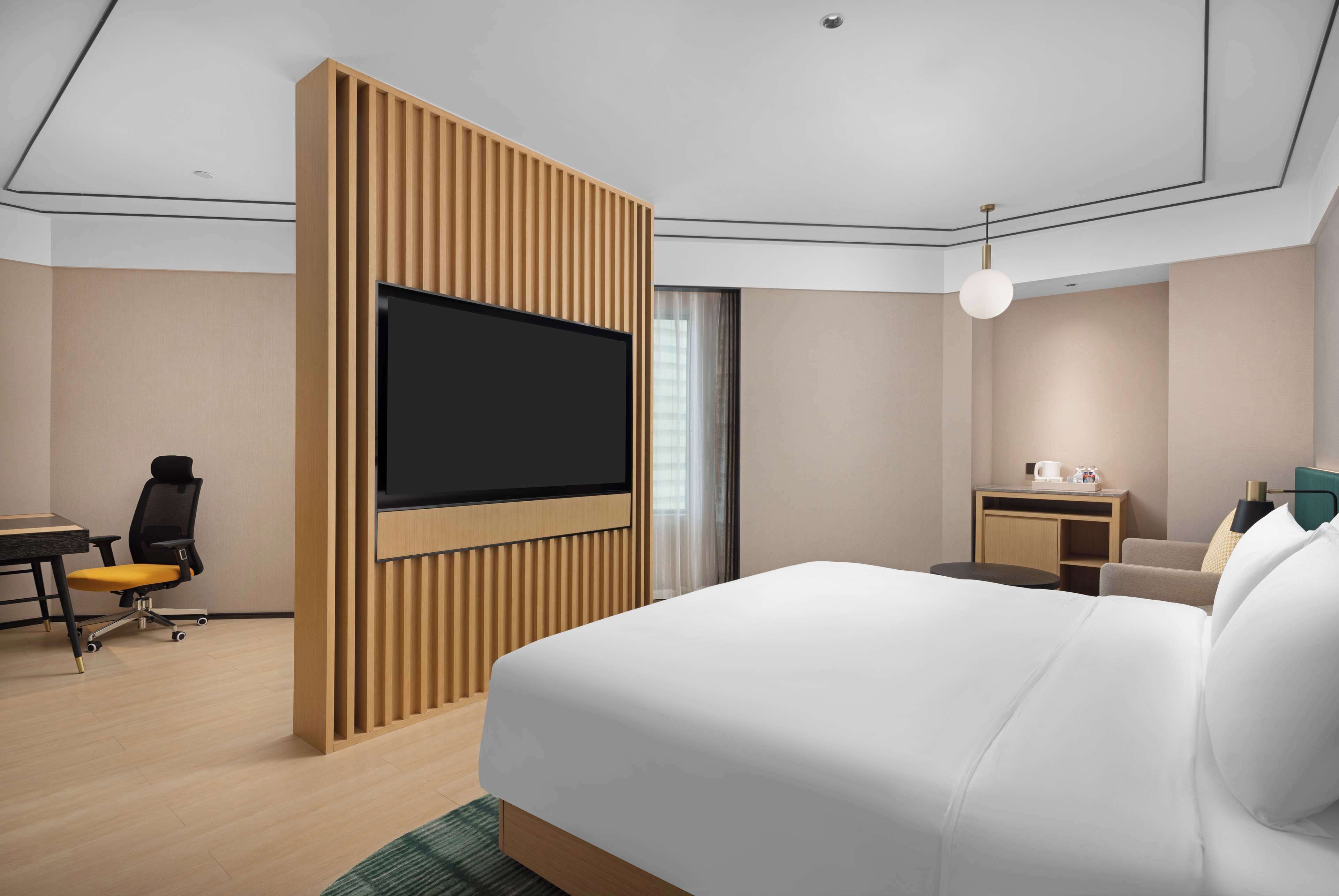 Quarto de hotel moderno com uma cama grande, uma TV de tela plana fixada em um painel de madeira, uma escrivaninha com uma cadeira e uma pequena mesa lateral.