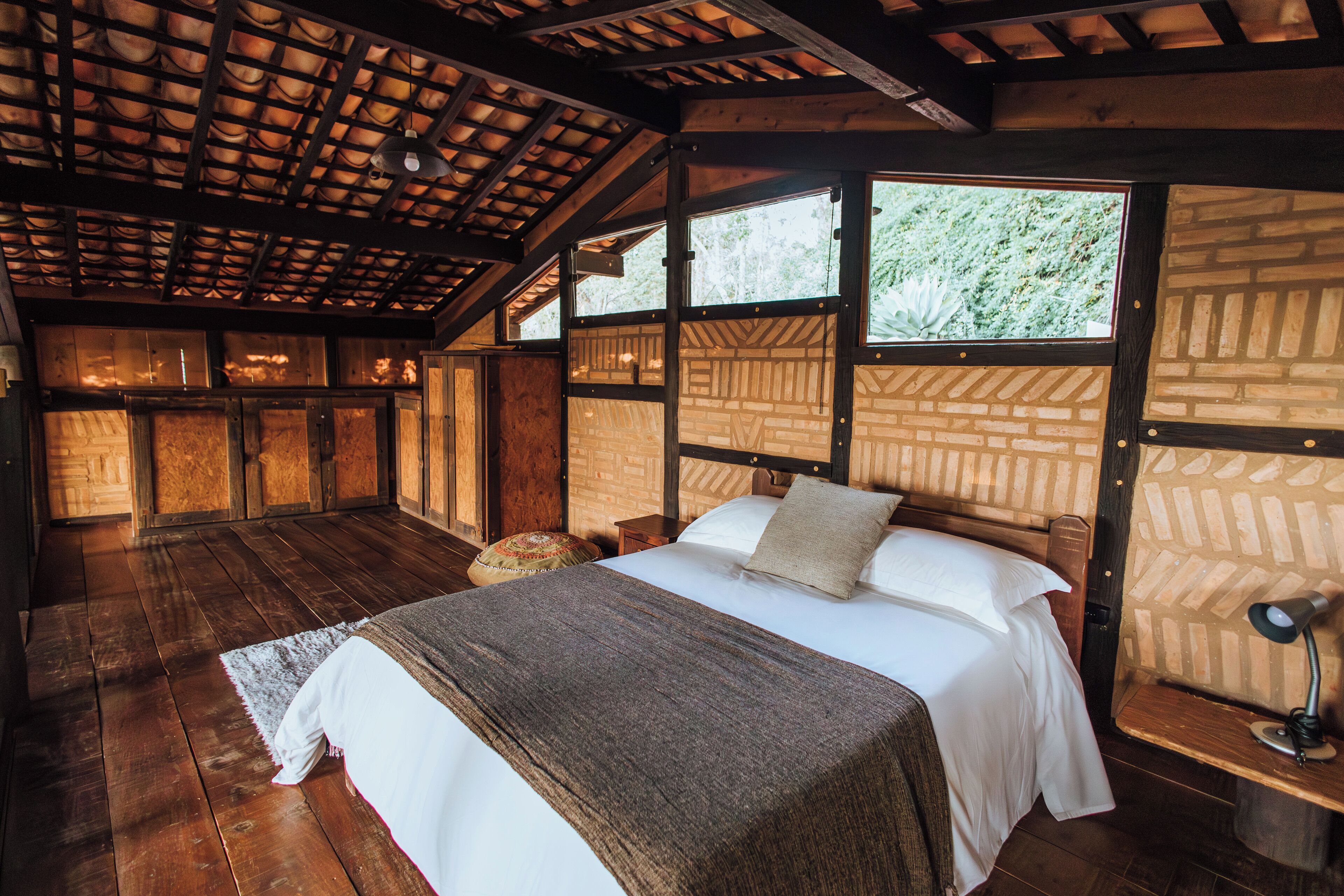Quarto de madeira com cama, mesa de cabeceira e janela com vista para árvores.