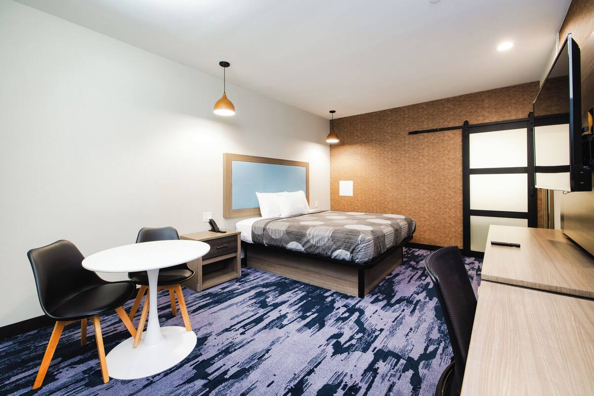 Quarto de hotel com cama, escrivaninha, cadeira, mesinha e televisão.