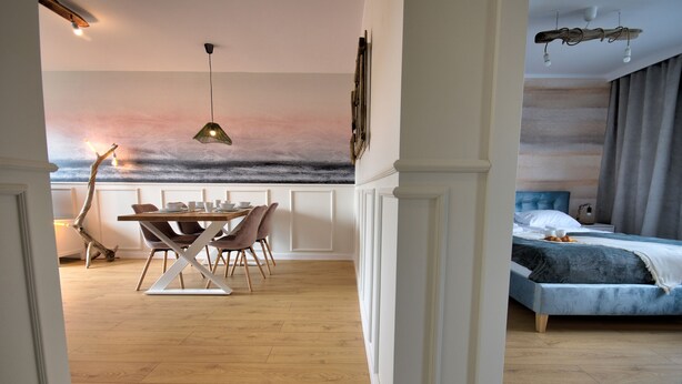 Quarto de hotel moderno com piso de madeira, área de jantar com mesa e cadeiras, cama com cabeceira azul e vista para o mar por uma grande janela.