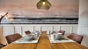 Uma área de refeições com uma mesa de madeira posta para quatro pessoas, pratos, tigelas e xícaras brancas, e um mural texturizado no fundo.
