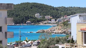 Uma vila costeira com edifícios dando para um mar azul límpido, um litoral rochoso e uma colina arborizada ao fundo.