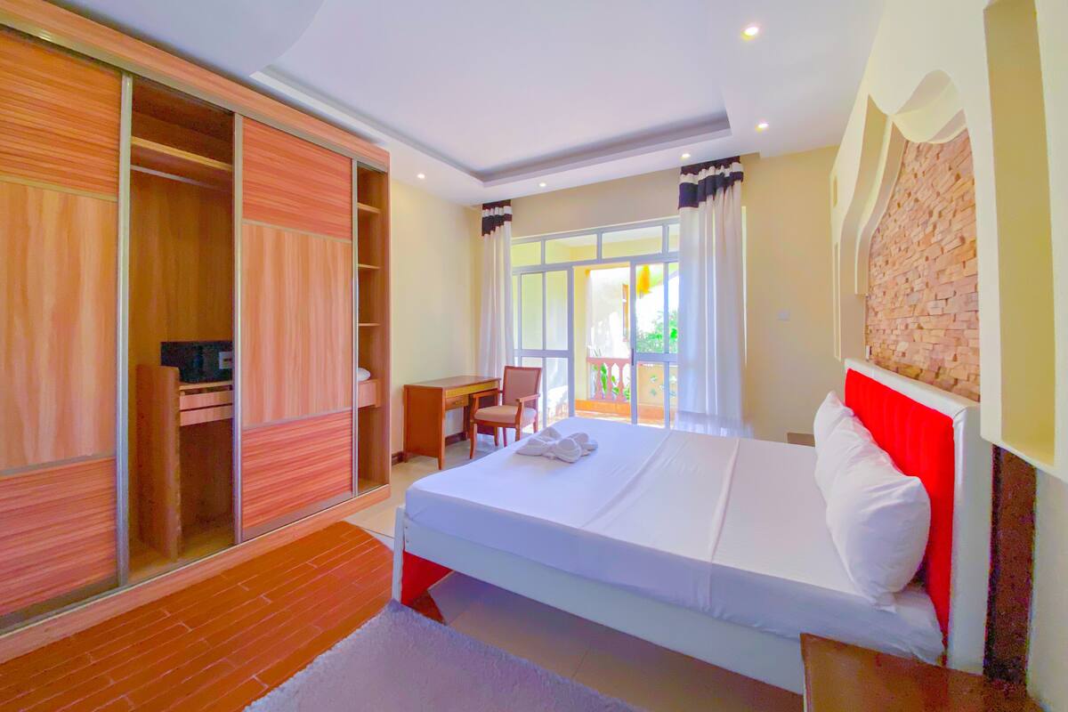 Quarto de hotel com uma cama, uma escrivaninha e uma cadeira. Possui um guarda-roupa de portas deslizantes e vista para o exterior.
