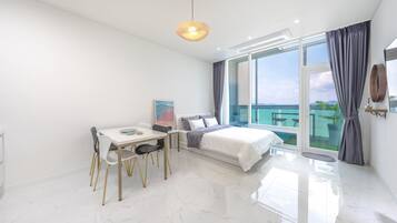 Quarto moderno com cama grande, mesa de jantar e vista para o mar através de amplas janelas.