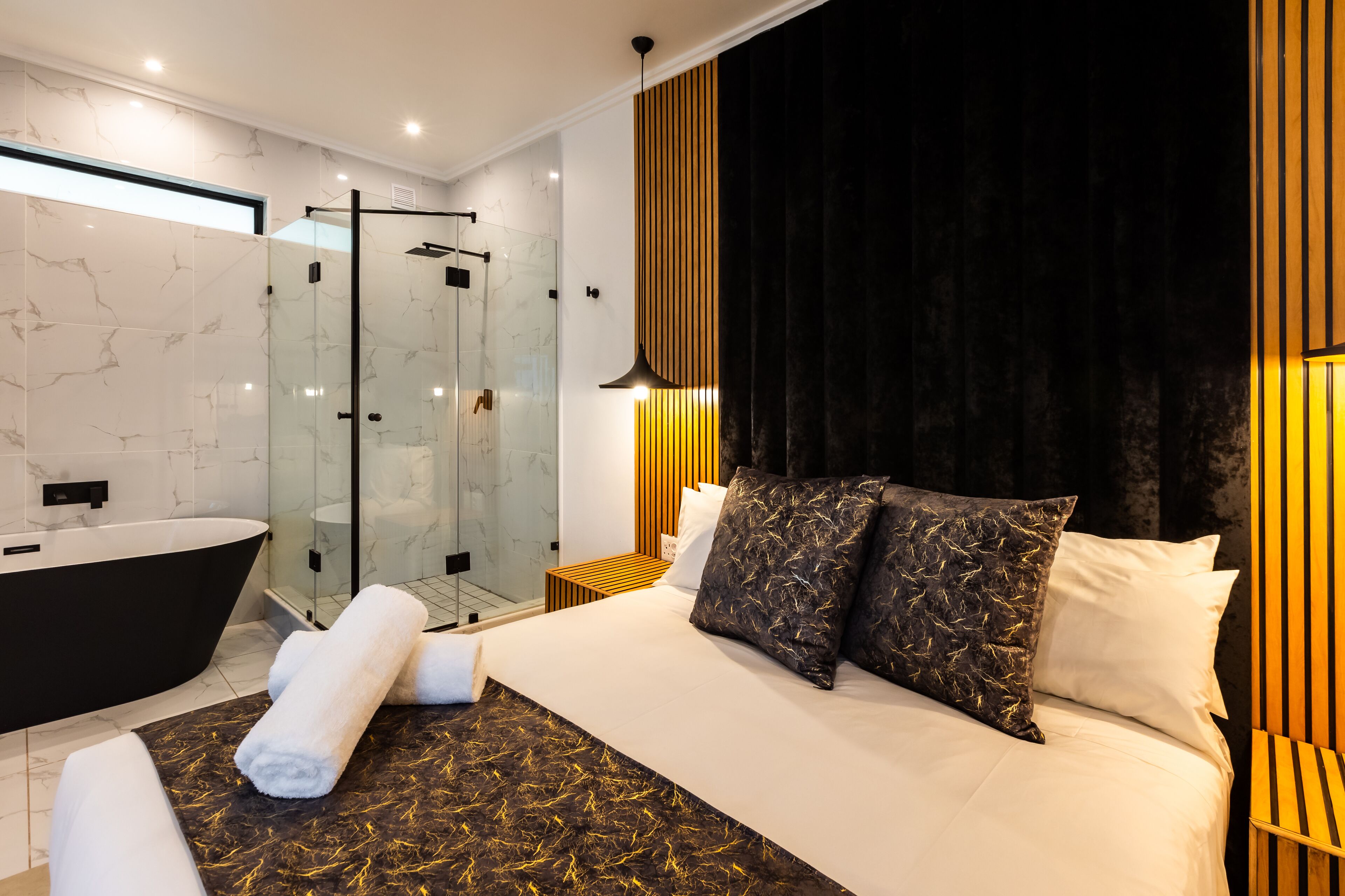 Quarto de hotel moderno com uma cama grande, um chuveiro com paredes de vidro, uma banheira preta e uma cabeceira de madeira.