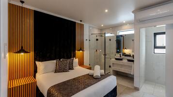 Quarto de hotel moderno com uma cama grande, box de vidro para o chuveiro, toucador com espelho e vista para a cidade.