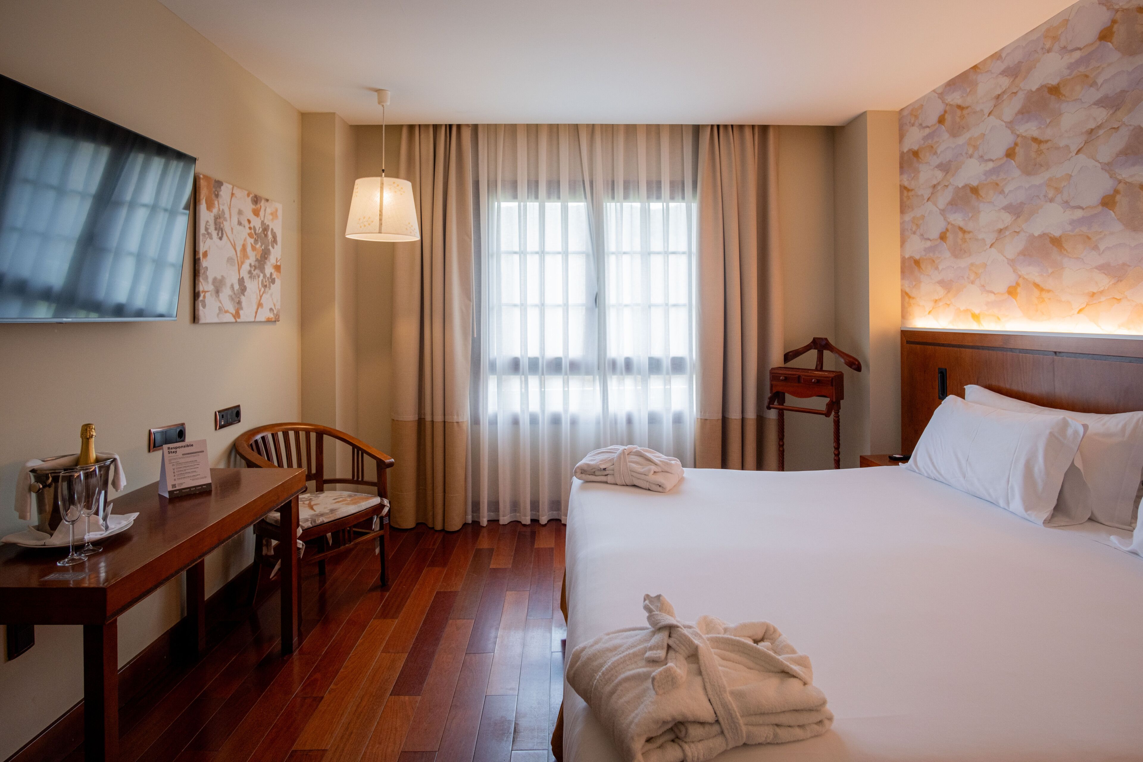 Quarto de hotel com uma cama grande, uma escrivaninha de madeira com uma garrafa e copos, uma cadeira, uma televisão na parede e uma janela com cortinas.