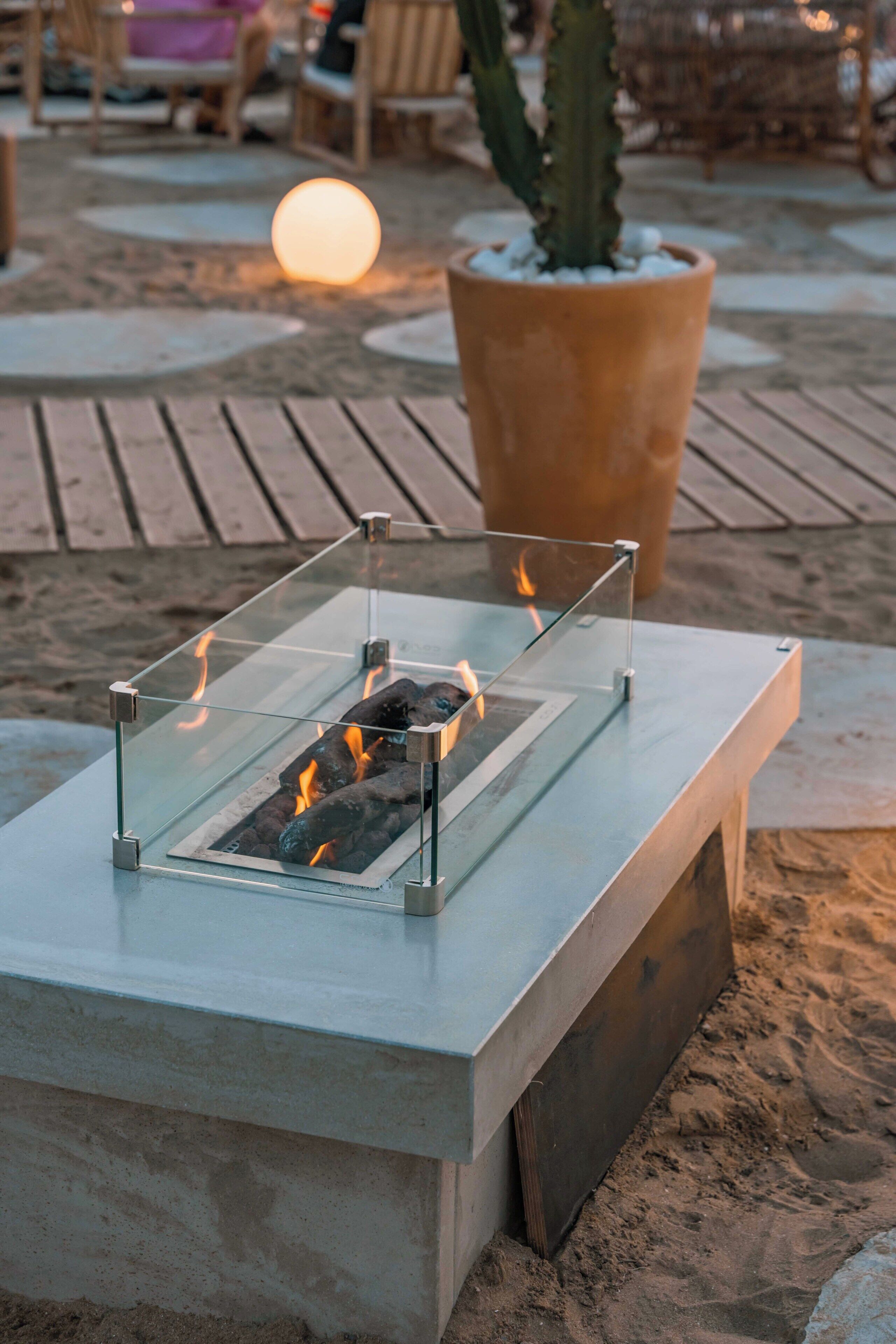 Uma lareira com mesa de vidro e brasas acesas, sobre base de concreto, rodeada por uma área externa arenosa com um cacto grande em vaso e uma luz redonda iluminada ao fundo.