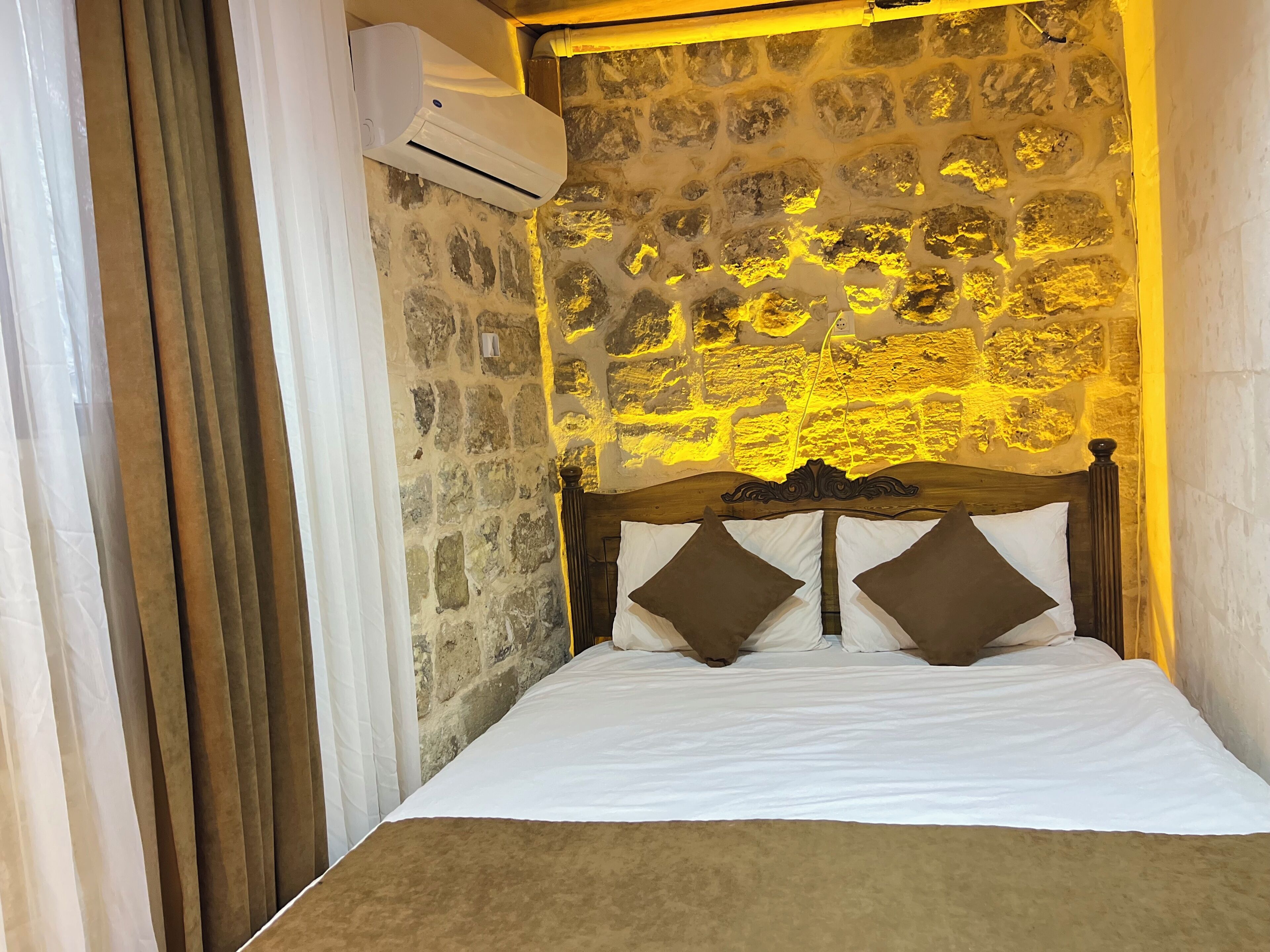 Quarto com parede de pedra, cama com roupa de cama branca e ar condicionado fixado na parede.