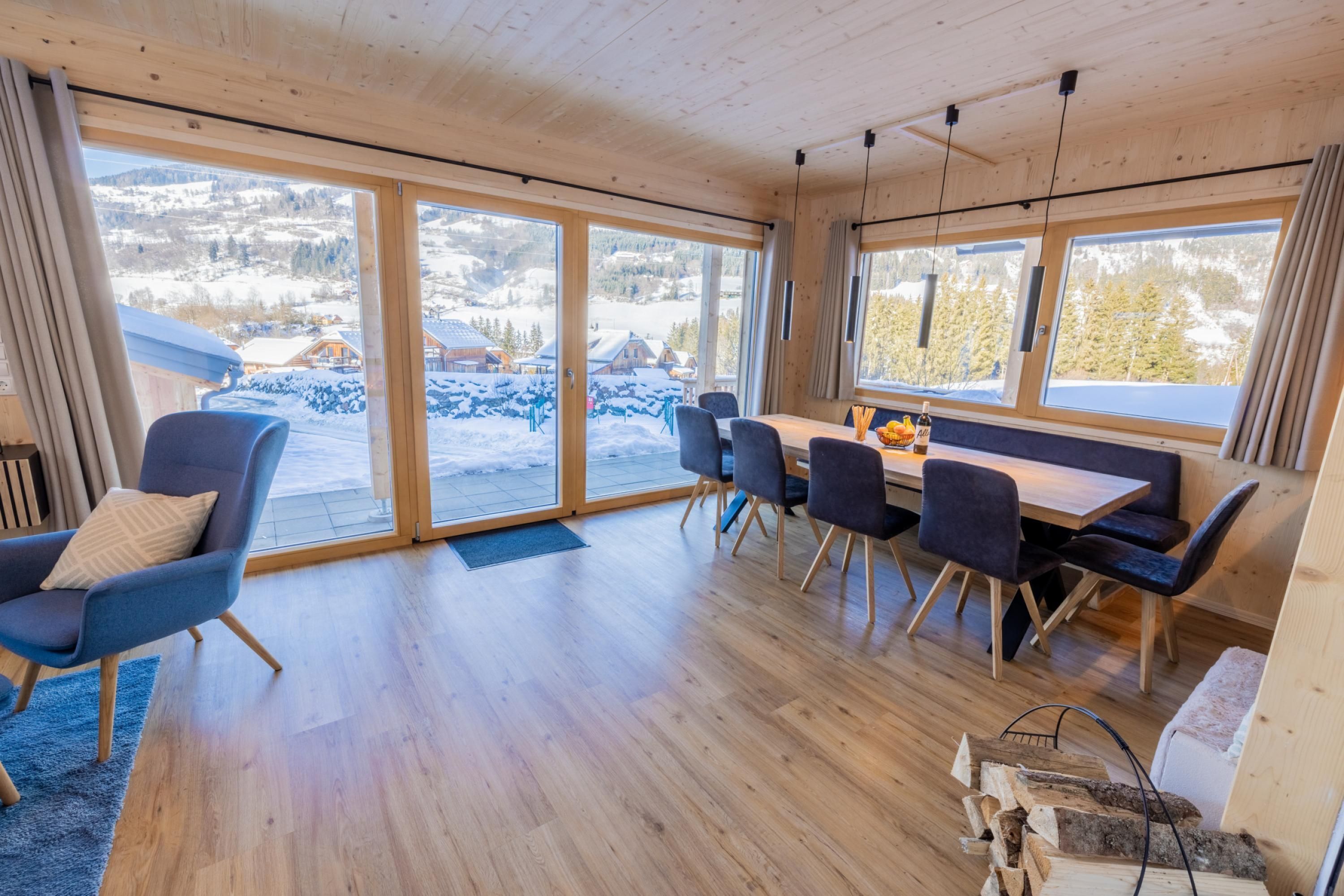 Um interior moderno com piso de madeira, sala de jantar com mesa e cadeiras, e grandes janelas com vista para uma paisagem nevada.