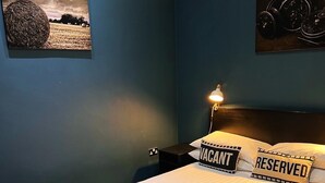 Quarto com cama, duas almofadas, mesa de cabeceira, abajur e quadro na parede.