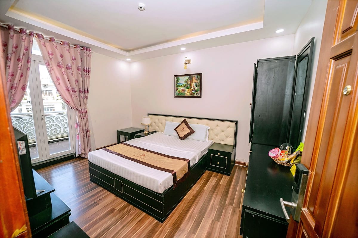 Quarto de hotel com cama, mesas de cabeceira, guarda-roupa, um quadro na parede e varanda com cortinas.
