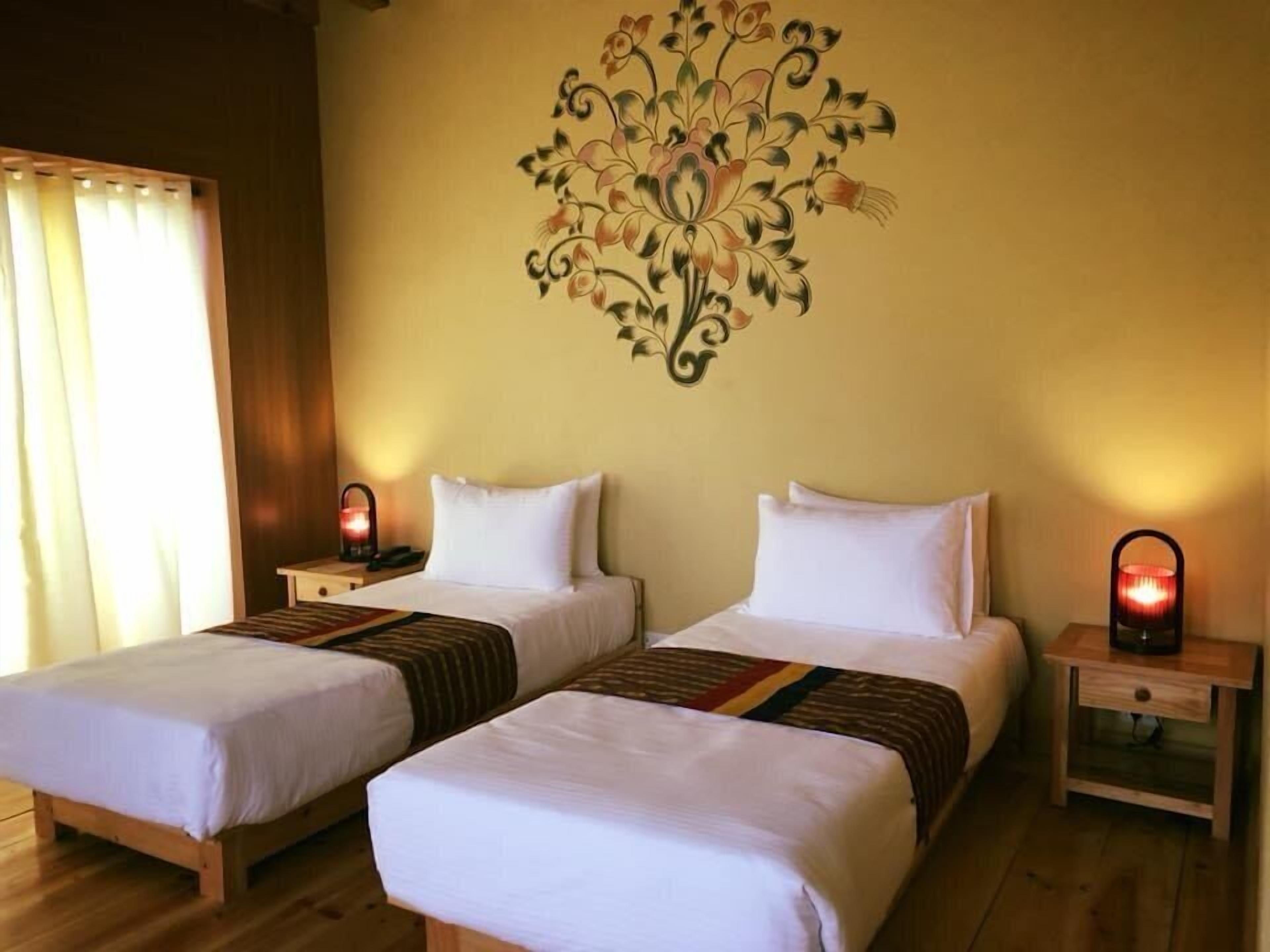 Quarto de hotel com duas camas de solteiro, um detalhe decorativo na parede, luminárias de cabeceira e uma janela com cortinas.