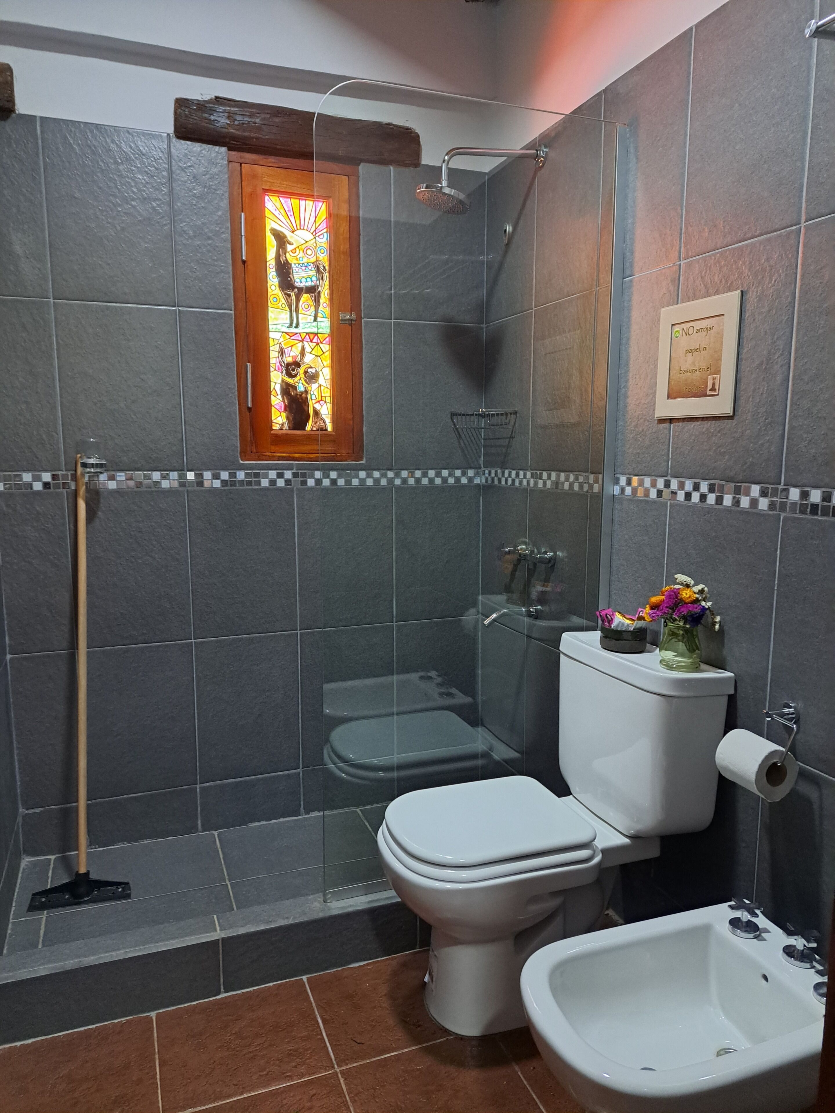 Casa de banho com cabine de duche em vidro, sanita, bidé e janela decorativa com um desenho colorido.