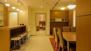 Quarto de hotel moderno com uma receção em madeira, área de refeições com mesa e cadeiras, e uma kitchenette ao fundo.