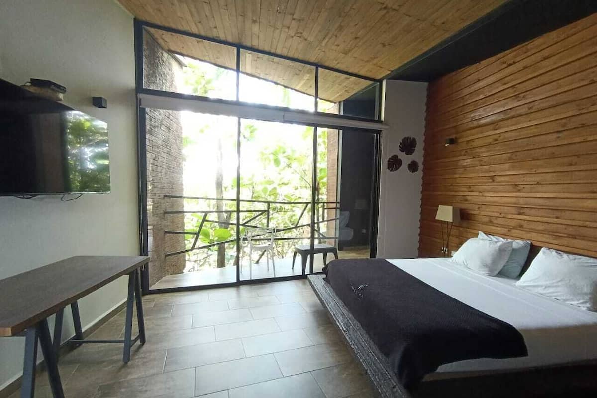 Quarto com cama, um banco, televisão e varanda com vista para o verde.
