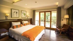 Quarto de hotel com uma cama grande, cabeceira de madeira, varanda com cadeiras e quadros nas paredes.