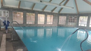Piscina interior com água cristalina, grandes janelas e teto alto com vigas de madeira.