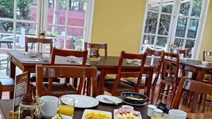 Uma área de refeições com mesas e cadeiras de madeira, itens de café da manhã como pão, frutas e ovos, e janelas amplas com vista para vegetação.