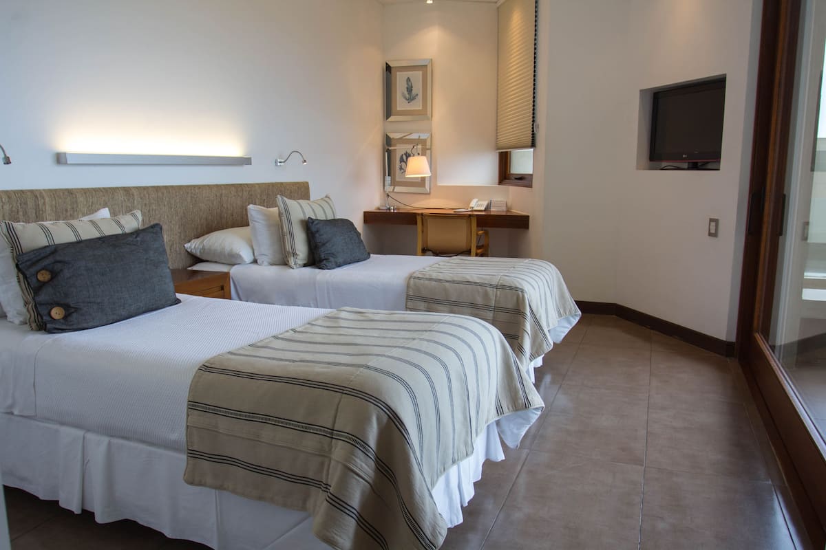 Quarto de hotel com duas camas, uma escrivaninha, uma cadeira, uma TV e uma mesinha de cabeceira.