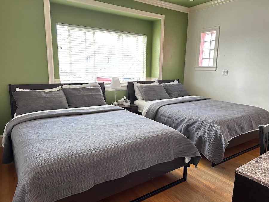 Quarto com duas camas, janela com persianas, mesa de cabeceira com abajur e uma pequena mesa com uma cadeira.