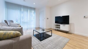 Uma sala de estar moderna com um sofá, uma mesa de centro de vidro, uma TV de tela plana em um suporte branco e uma janela grande com persianas.