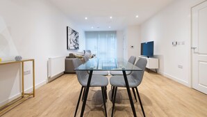 Uma sala de estar moderna com uma mesa de jantar de vidro, cadeiras cinzas, um sofá, uma TV e uma janela grande com cortinas leves.