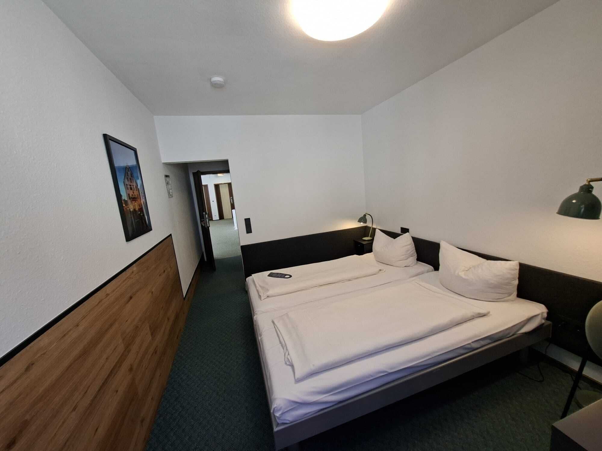 Quarto de hotel com cama, mesa de cabeceira, luminária e um quadro na parede.