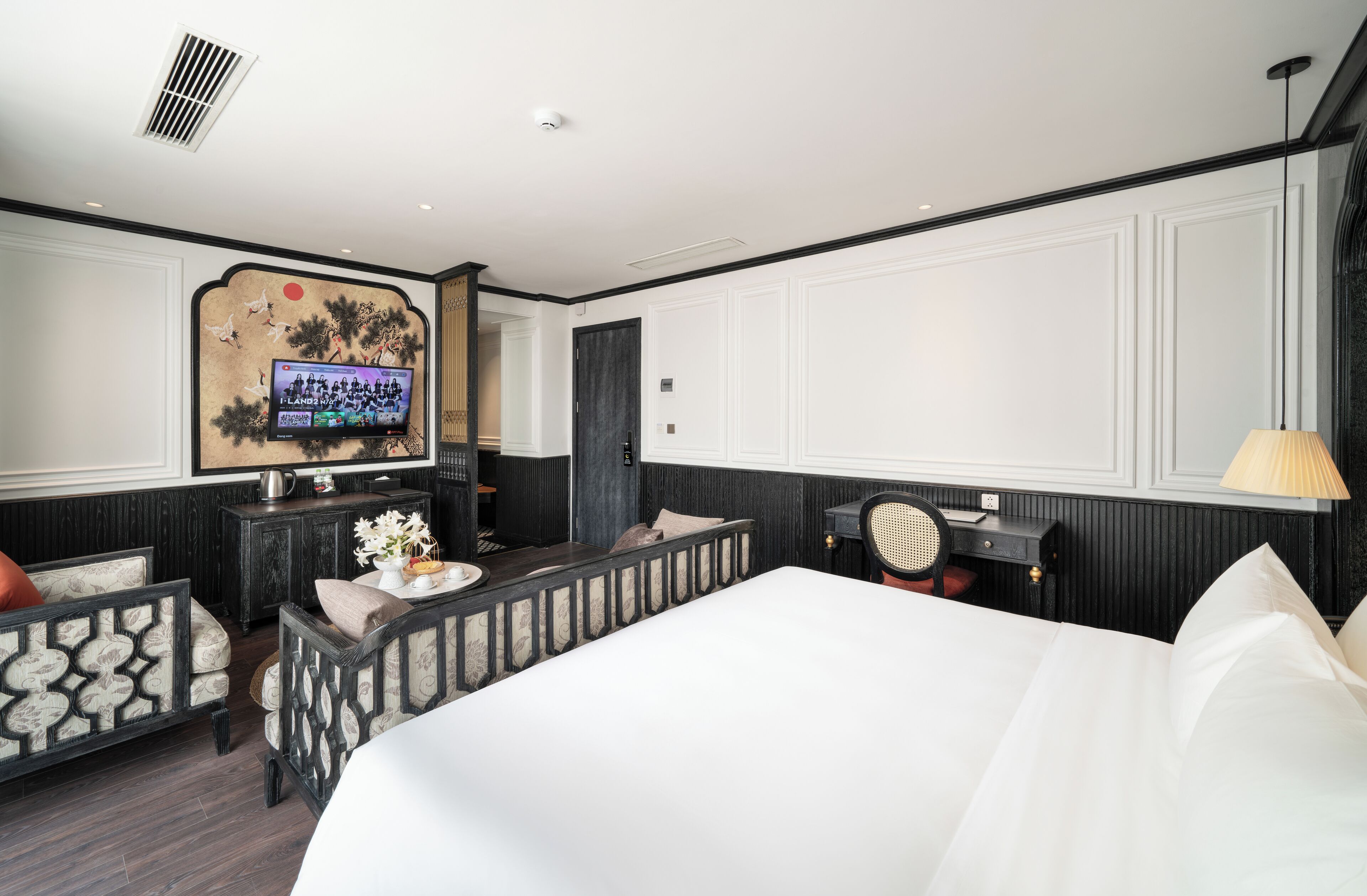 Quarto de hotel com uma cama grande, televisão na parede, escrivaninha com luminária e área de estar com cadeiras e mesa.