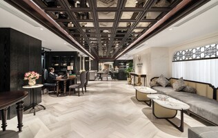 Um hall de hotel moderno com padrão de piso em formato de losango, uma área de estar central com um sofá e poltronas, e uma área de jantar com mesa e cadeiras.
