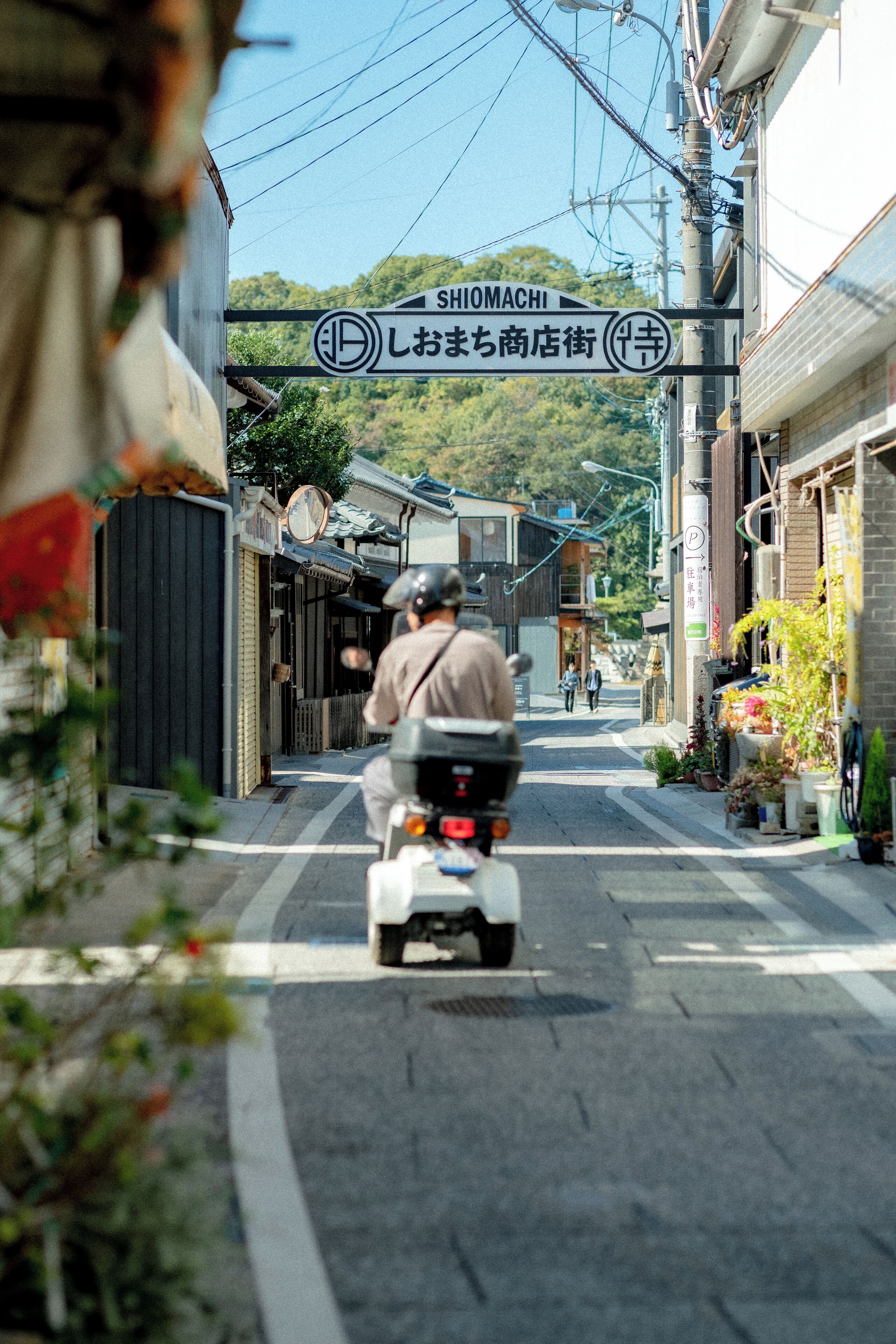 Cena de rua com uma moto em primeiro plano, uma placa indicando 'SHIOMACHI' e 'Shomachi Shopping Street' ao fundo, e edifícios ao longo da rua.