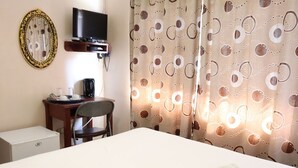 Quarto de hotel com cama, escrivaninha, cadeira, televisão, máquina de café e pequeno frigorífico.