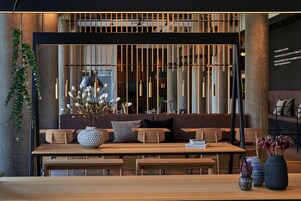 Um interior moderno com uma mesa de jantar de madeira, bancos e um sofá com almofadas. Há vasos decorativos na mesa e uma planta pendente à esquerda.