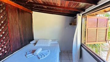 Uma cama de solteiro com roupa de cama e toalhas brancas, teto de madeira e janela com persianas de madeira.