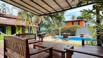 Um deck de madeira com uma cadeira suspensa, uma piscina e um edifício com telhado cerâmico ao fundo.