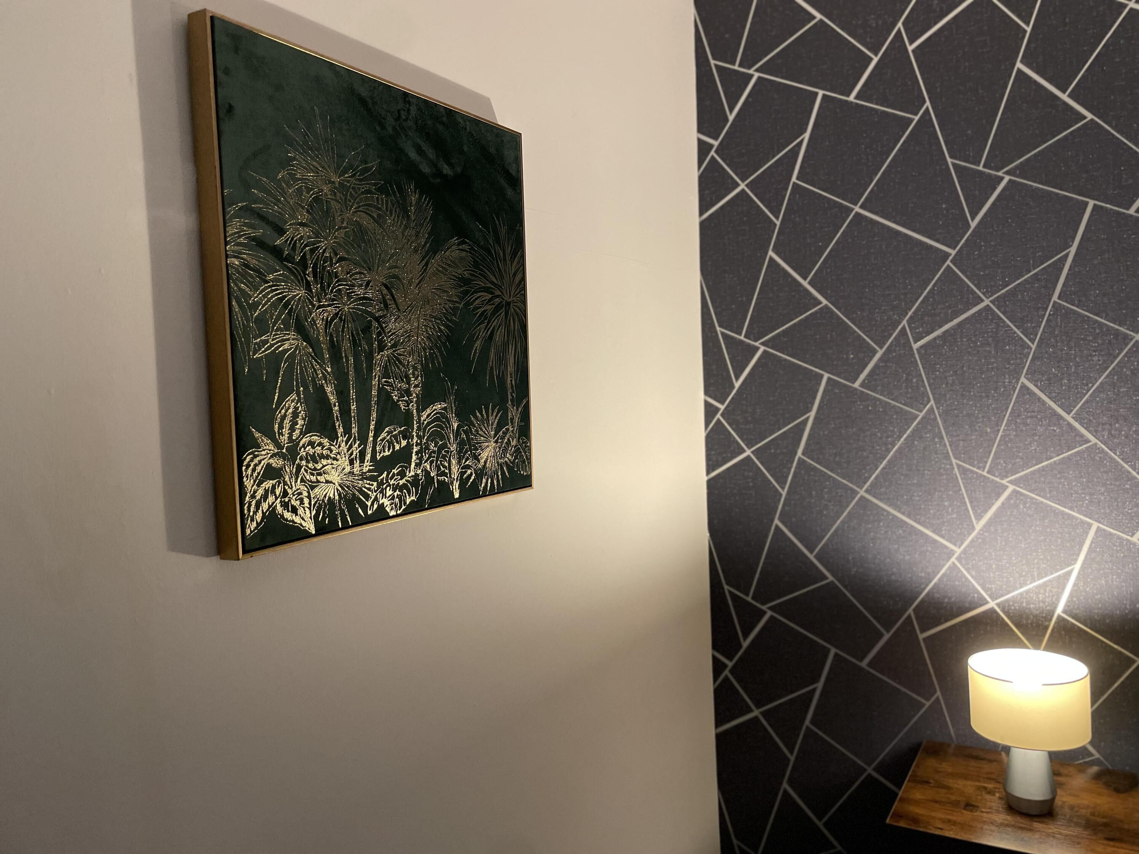 Uma obra de arte emoldurada com tema tropical, parede com padrão geométrico e uma luminária de mesa.